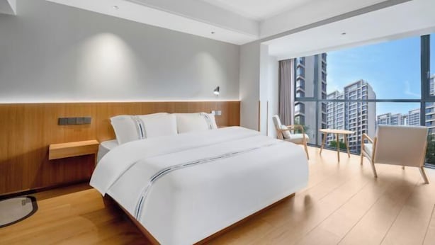 Quarto de hotel moderno com uma cama grande, uma mesa pequena e vista da cidade através de janelas amplas.