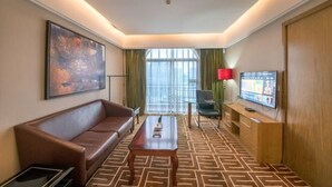 Quarto de hotel com um sofá de couro marrom, uma mesa de centro de madeira, uma televisão sobre um suporte de madeira, uma janela grande com cortinas e um quadro na parede.