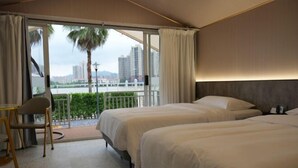 Quarto de hotel com duas camas, uma varanda com vista para palmeiras e edifícios, e uma pequena mesa com uma cadeira.
