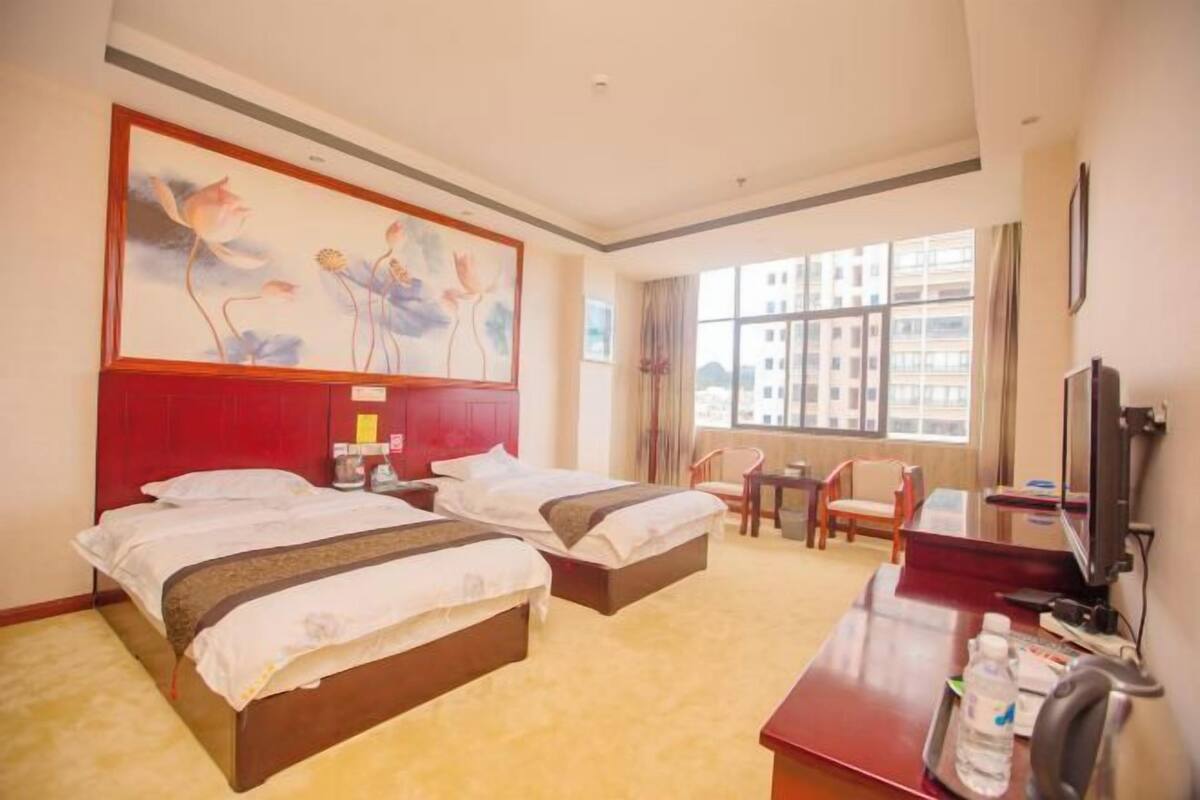 Quarto de hotel com duas camas, uma escrivaninha, uma cadeira, televisão e uma janela grande com vista para edifícios.