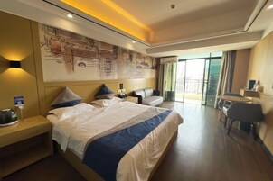 Quarto de hotel moderno com uma cama grande, área de estar com sofá e poltronas, uma mesinha e uma janela ampla com vista para a cidade.