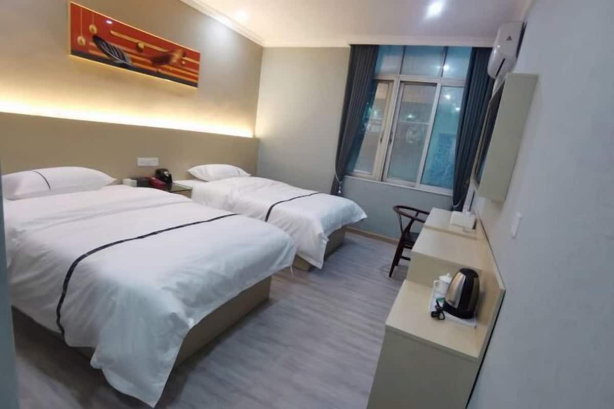 Quarto de hotel com duas camas, uma pequena mesa com chaleira, janela com cortinas e ar condicionado fixado na parede.