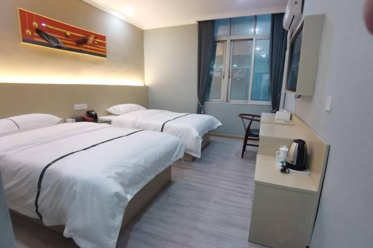 Quarto de hotel com duas camas, uma secretária pequena, uma cadeira, janela com cortinas e ar condicionado fixado na parede.