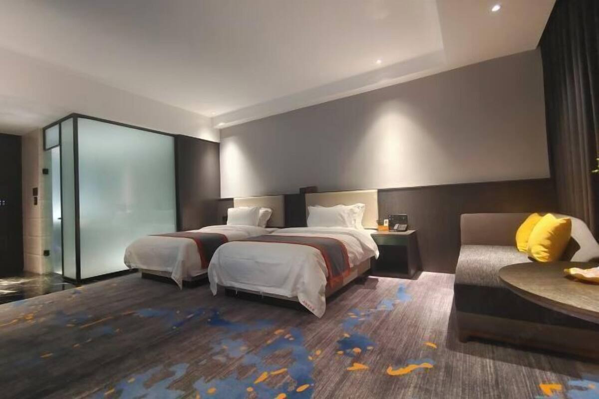 Quarto de hotel moderno com duas camas, um sofá, uma escrivaninha e um painel de vidro.