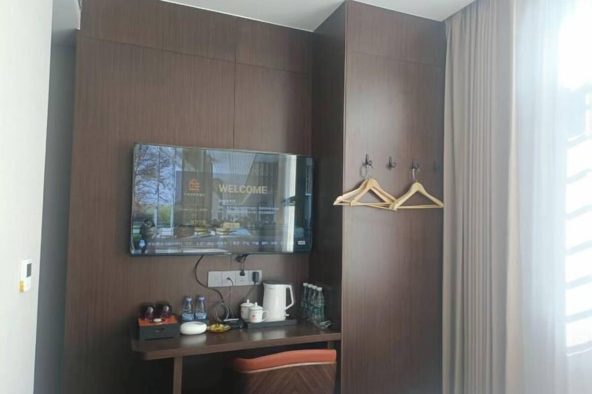 Quarto de hotel com televisão na parede exibindo uma tela de boas-vindas, escrivaninha com cafeteira, garrafas de água e uma pequena planta.