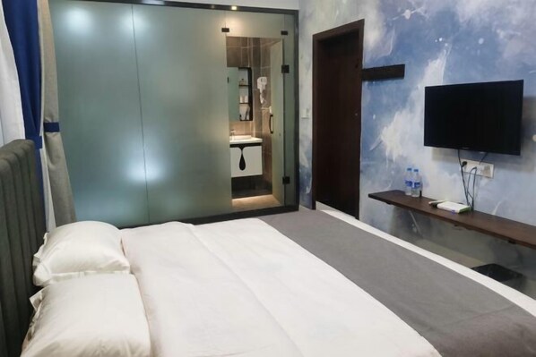 Quarto com cama, televisão na parede, pequena prateleira com objetos e porta de acesso a um banheiro.
