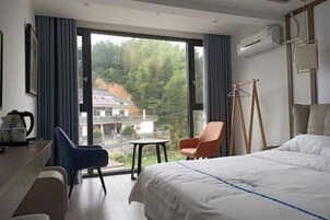 Quarto de hotel com cama, escrivaninha, cadeira, mesa pequena e vista para uma área arborizada.