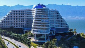 Um hotel moderno com design arquitetônico único, apresentando uma cúpula central e várias varandas, situado perto de um corpo d'água com montanhas ao fundo.