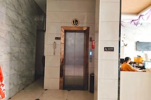 Um elevador com uma placa acima, um cesto de lixo ao lado e uma receção com uma pessoa sentada atrás.