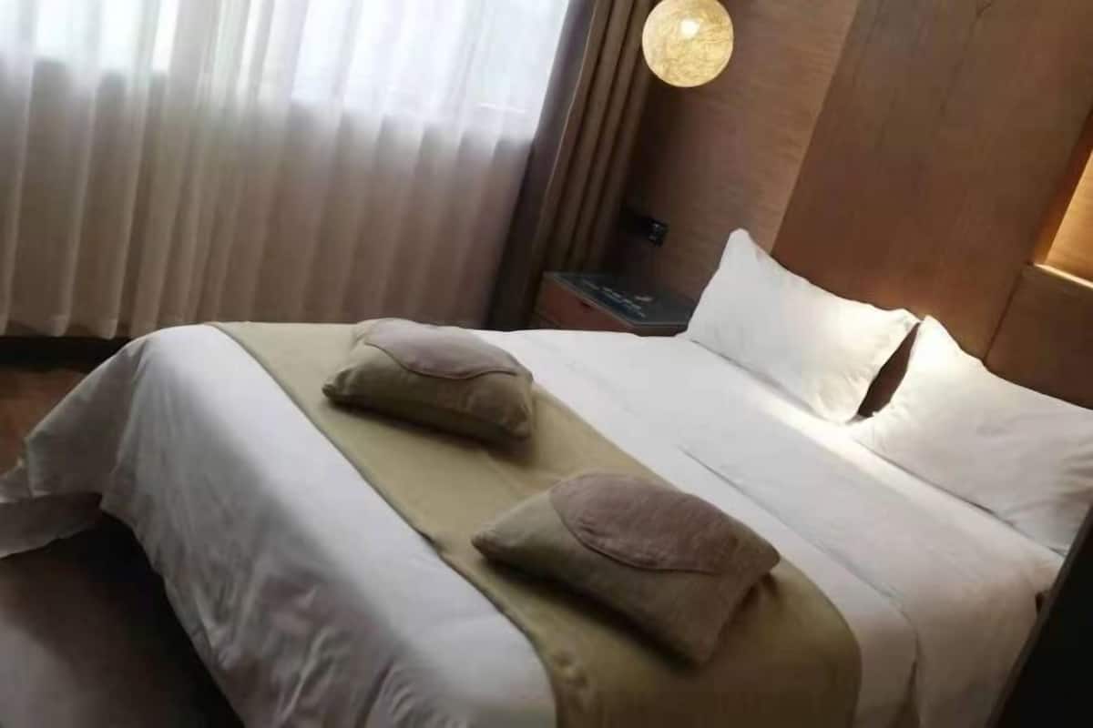 Quarto de hotel com uma cama bem arrumada, uma mesa de cabeceira com abajur e uma janela com cortinas.