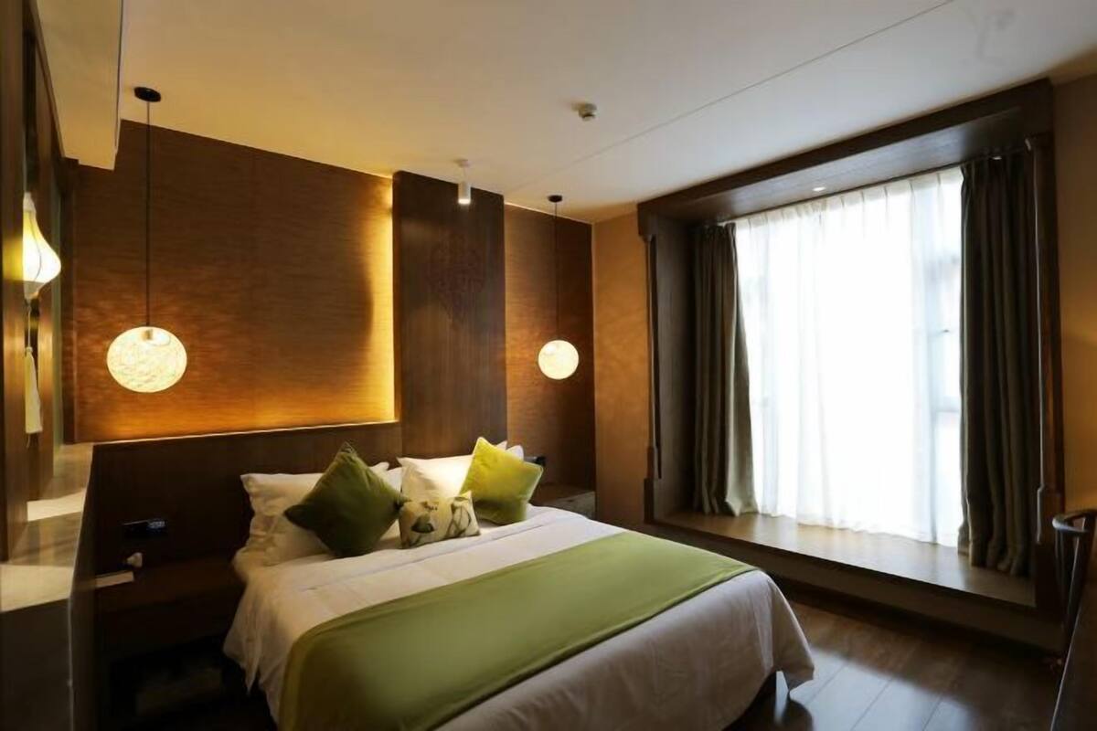 Quarto de hotel com uma cama grande, duas luminárias de cabeceira, uma janela com cortinas e uma cabeceira de madeira.