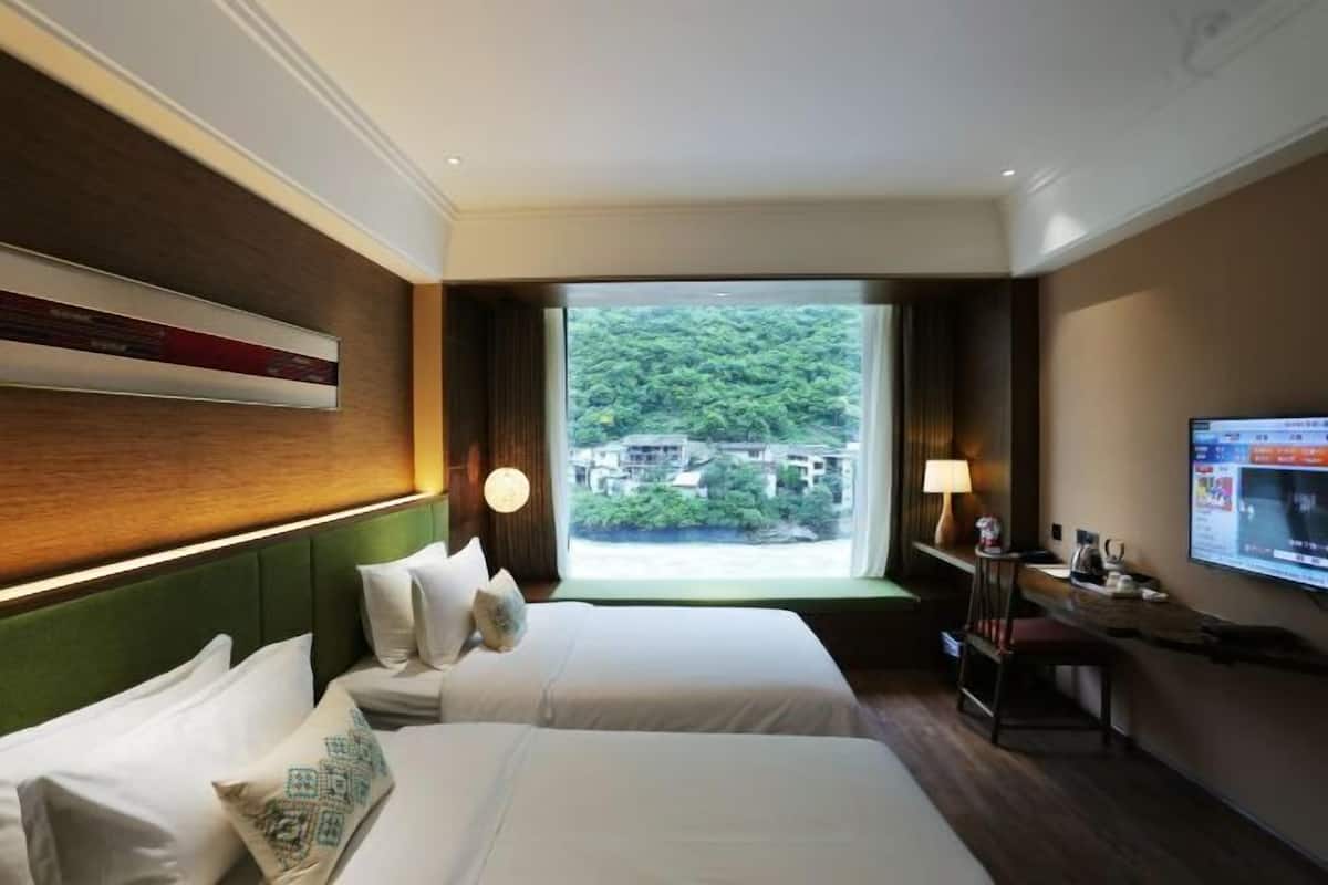 Quarto de hotel com duas camas, televisão, escrivaninha e janela com vista para áreas verdes e edifícios.