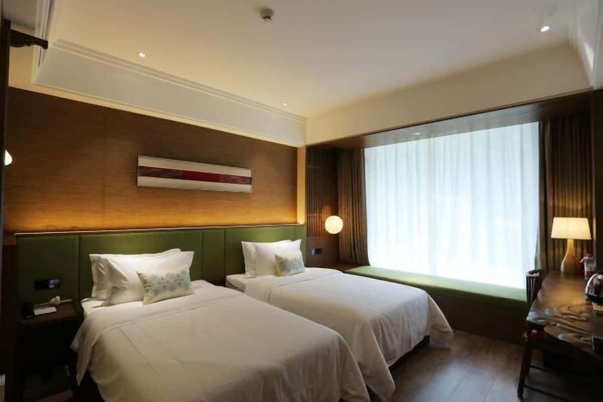 Quarto de hotel com duas camas, uma escrivaninha, uma cadeira, janela com cortinas e uma obra de arte na parede.