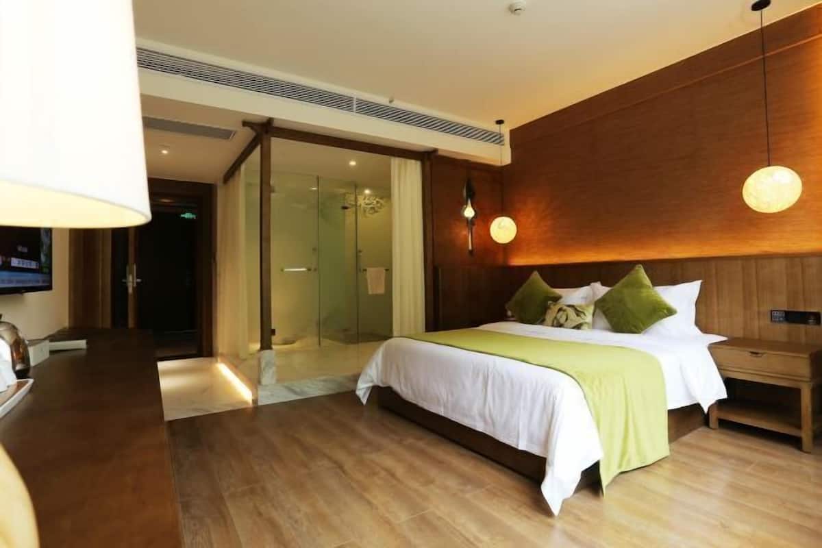 Quarto de hotel com cabeceira de madeira, uma cama grande com roupa de cama branca e almofadas verdes, uma mesa de cabeceira e uma TV montada na parede.