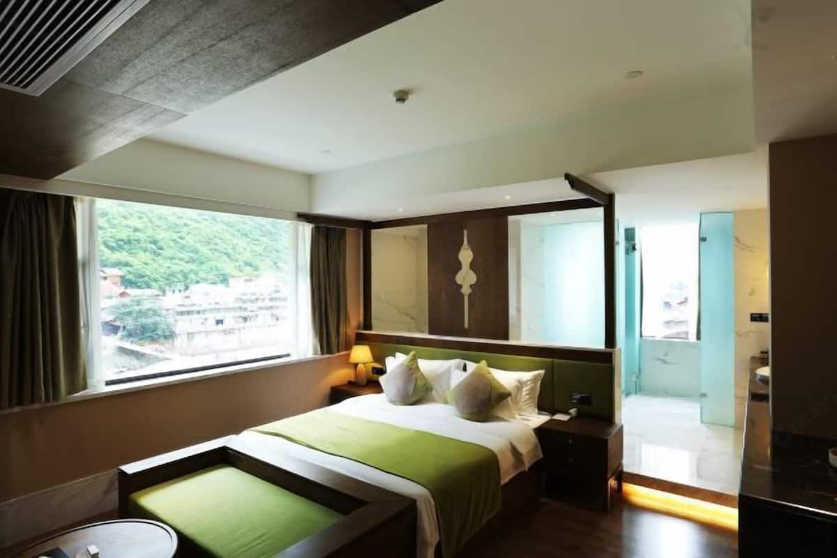 Quarto de hotel com cama, mesa de cabeceira, abajur e vista para o verde lá fora.