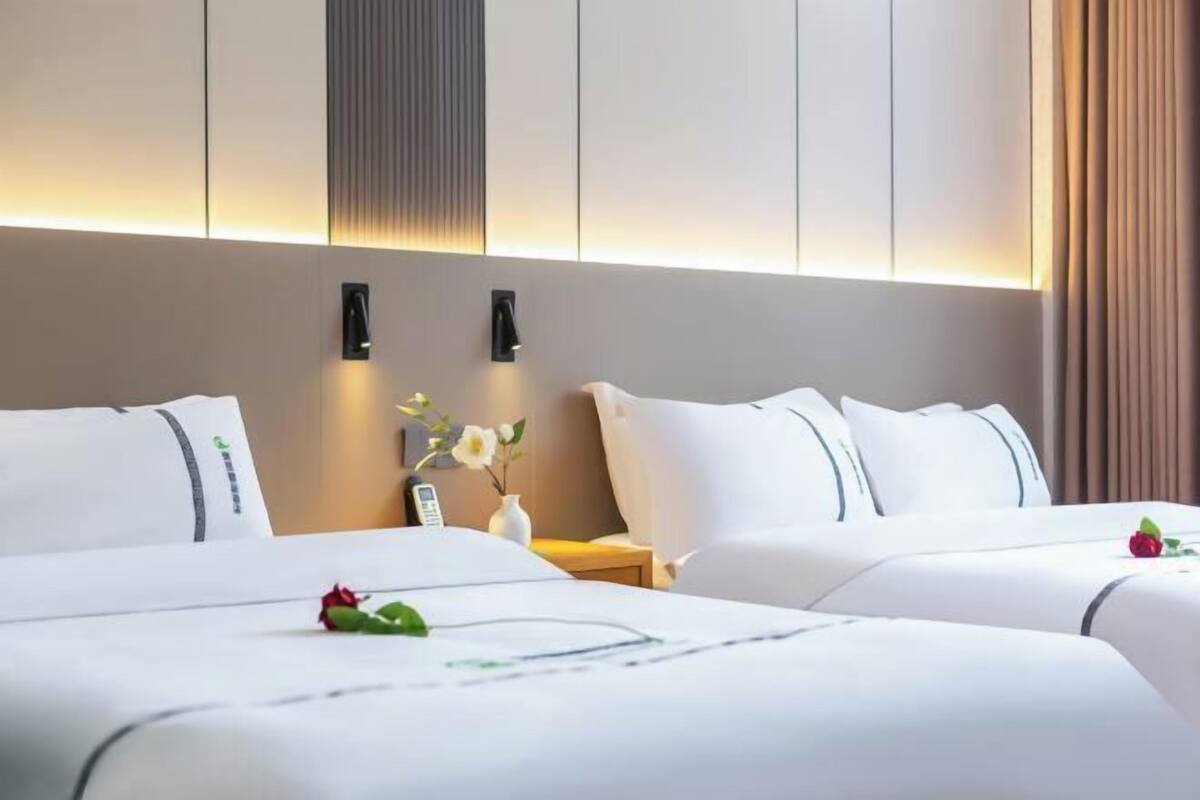 Quarto de hotel com duas camas, uma mesa de cabeceira com um vaso de flores e um candeeiro fixado na parede.