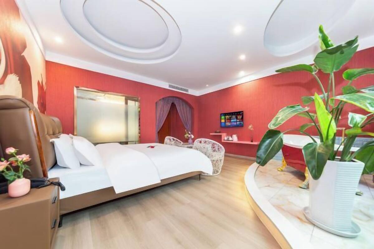 Quarto de hotel moderno com cama, área de estar, televisão e uma grande planta em vaso.