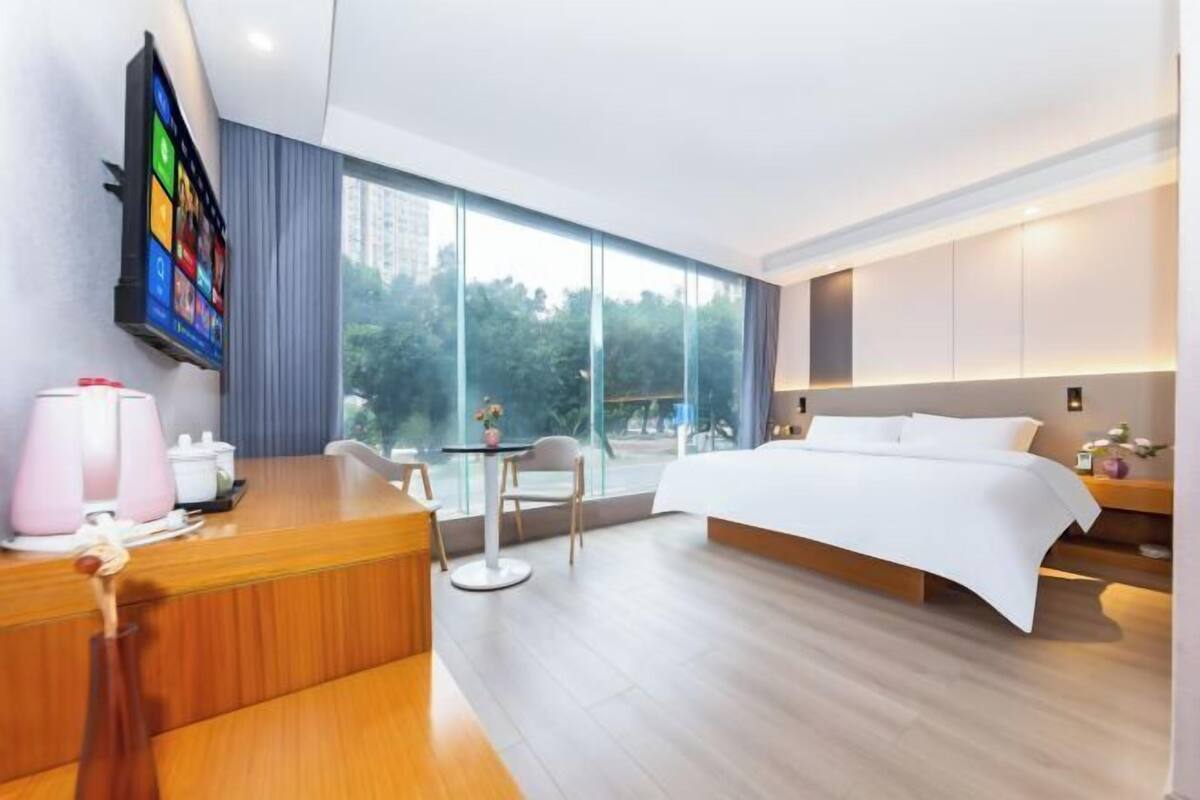 Quarto de hotel moderno com uma cama grande, uma escrivaninha com cadeira, uma TV na parede e uma janela ampla com vista para a cidade.