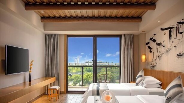 Quarto de hotel com duas camas, televisão, teto de madeira e uma janela grande com vista para a cidade.