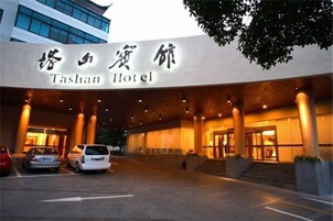 A entrada do Hotel Tashan com design moderno, destacando uma placa grande, uma área de acesso coberta e um estacionamento com vários carros.