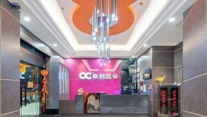 Uma área de recepção de hotel com design moderno, apresentando uma parede rosa com o nome do hotel, um balcão de recepção e um lustre decorativo.