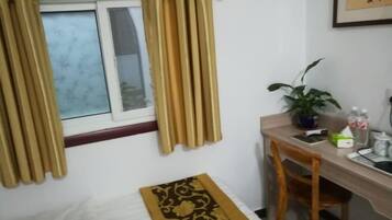 Quarto com cama, escrivaninha e cadeira, janela com cortinas, planta em vaso e pequena mesa com objetos.