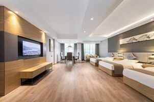 Quarto de hotel moderno com piso de madeira, uma televisão de tela plana grande fixada na parede, um sofá, uma cama e uma área de refeições com mesa e cadeiras.