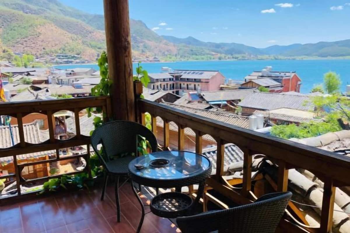 Uma varanda com vista para um lago e montanhas, equipada com cadeiras de vime e uma mesa de vidro.