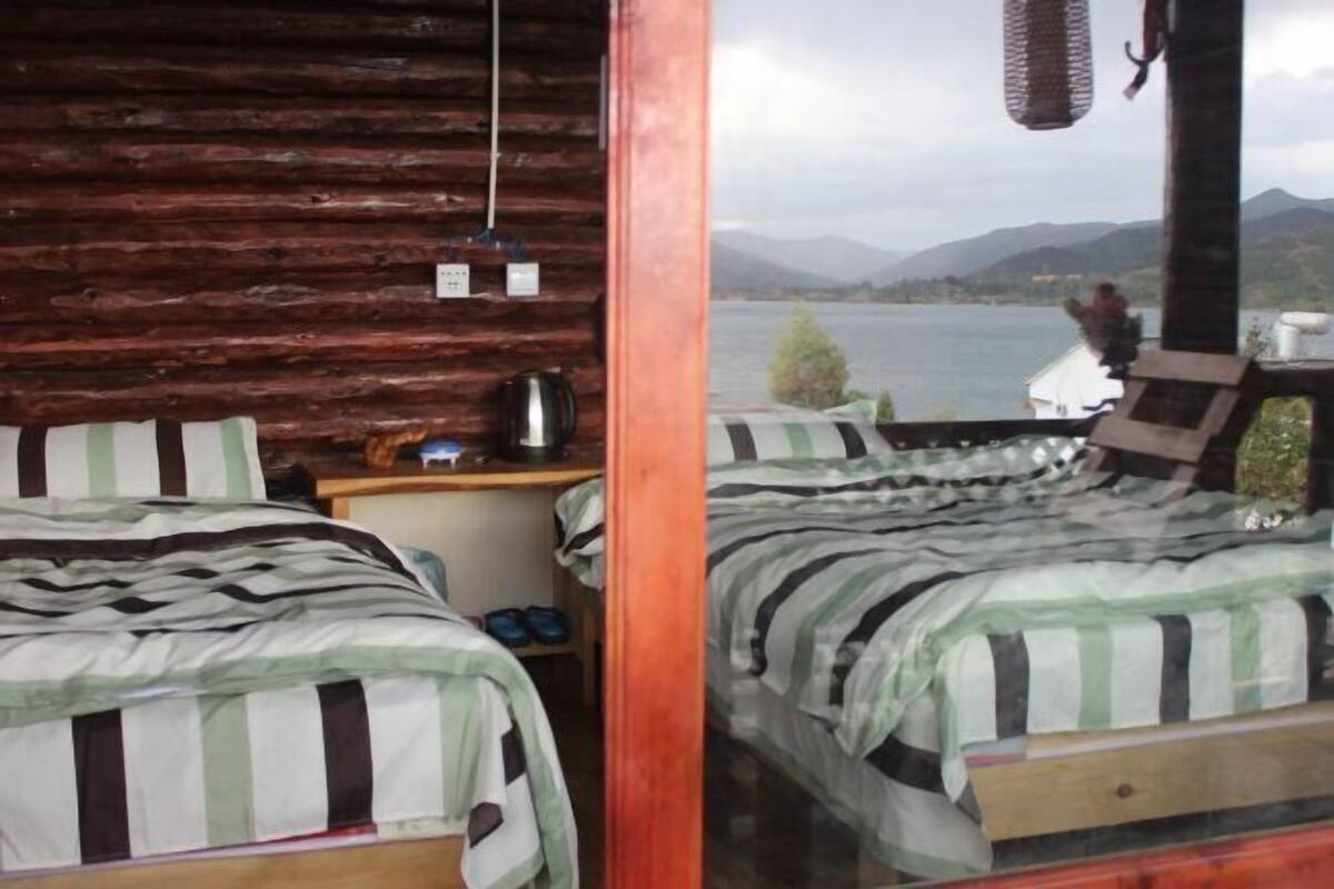 Uma cabana com duas camas, interior de madeira e vista para um lago e montanhas.
