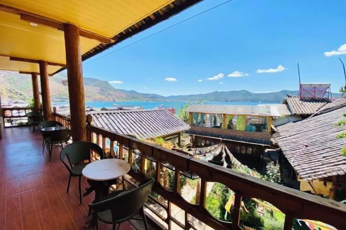 Um terraço com mesas e cadeiras, com vista para uma área costeira com edifícios tradicionais e montanhas ao fundo.