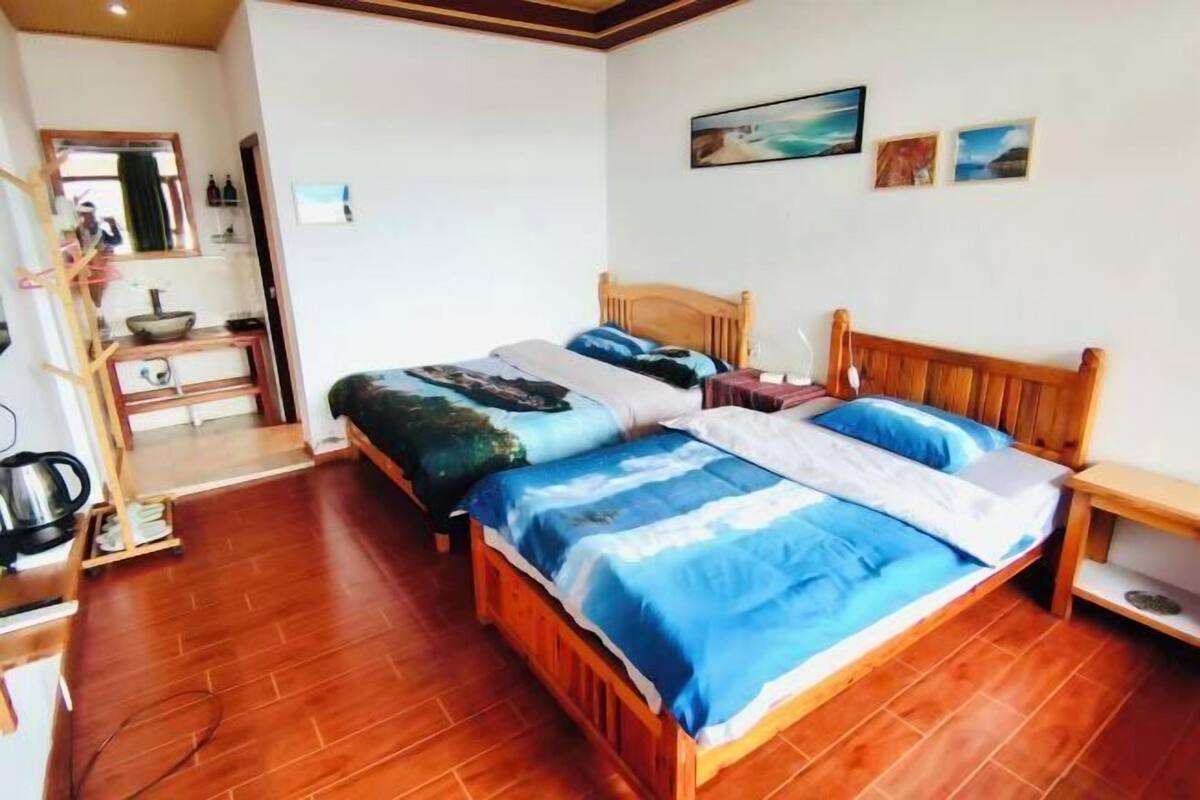 Quarto com duas camas de madeira, uma mesinha de cabeceira de madeira, uma pequena prateleira e um aparador de madeira com espelho.