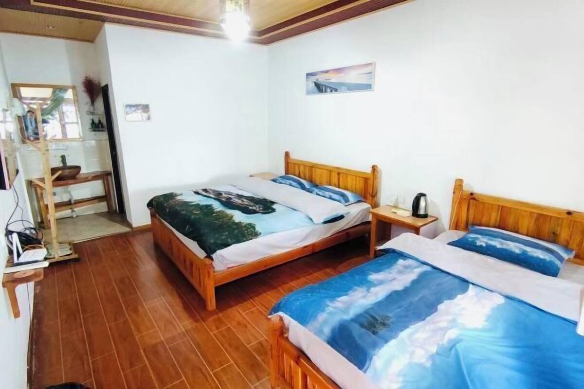 Quarto com duas camas de madeira, uma mesinha de cabeceira de madeira, uma pequena mesa e uma prateleira fixada na parede.