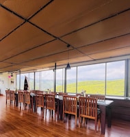 Uma sala de jantar ampla com cadeiras e mesas de madeira, janelas grandes que oferecem vista para uma paisagem verde, e um teto com padrão trançado.