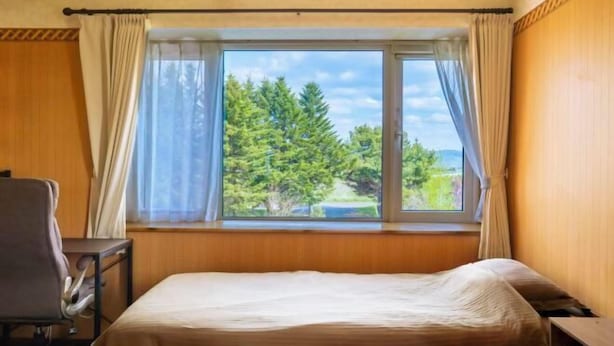 Quarto de hotel com cama, cadeira, escrivaninha e janela com cortinas.