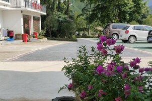 Um edifício com varanda, área de estacionamento com dois carros e canteiro de flores roxas.