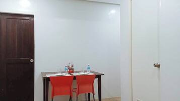 Um quarto pequeno com porta de madeira, parede branca e uma mesa com duas cadeiras vermelhas. Há garrafas de água sobre a mesa.