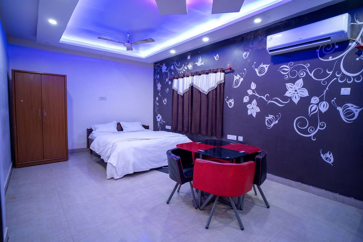 Quarto com cama, guarda-roupa, mesa e cadeiras, e parede com motivos florais.