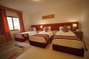 Quarto de hotel com três camas, uma janela grande com cortinas, uma cabeceira de madeira e um tapete estampado.