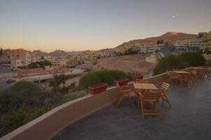 Um terraço ao ar livre com mesas e cadeiras de madeira, com vista para uma paisagem urbana com edifícios e uma montanha ao longe.