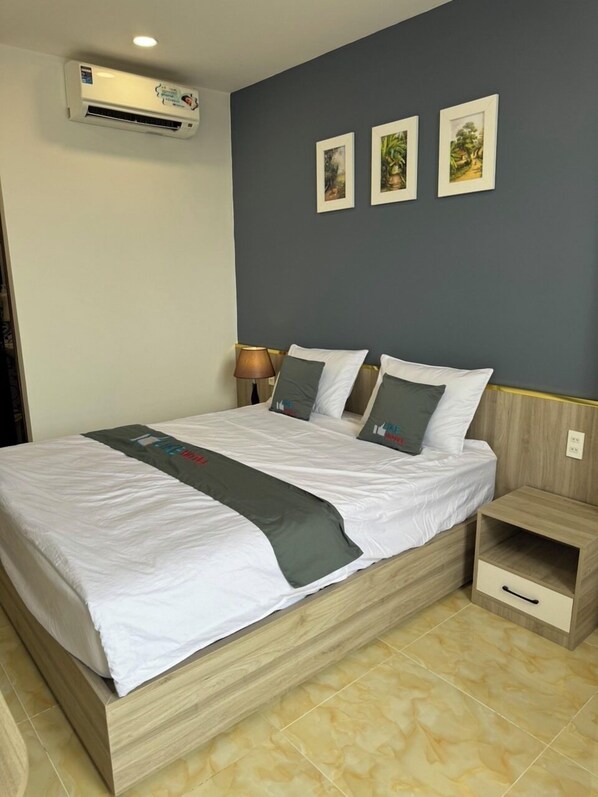 Cama bem arrumada com roupa de cama branca e detalhes em cinza, uma mesa de cabeceira com abajur, e três quadros na parede.