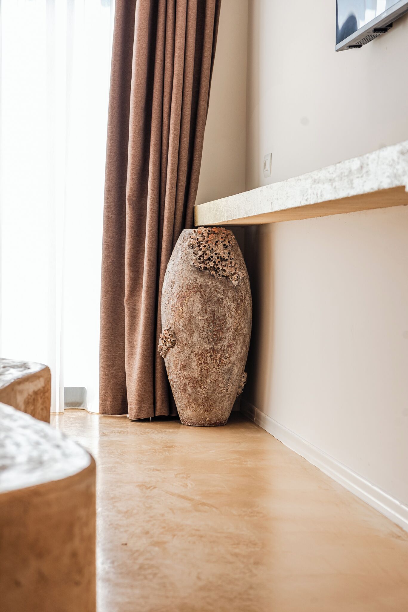 Canto de um quarto com um vaso de pedra texturizado, um banco de madeira e uma prateleira fixada na parede.