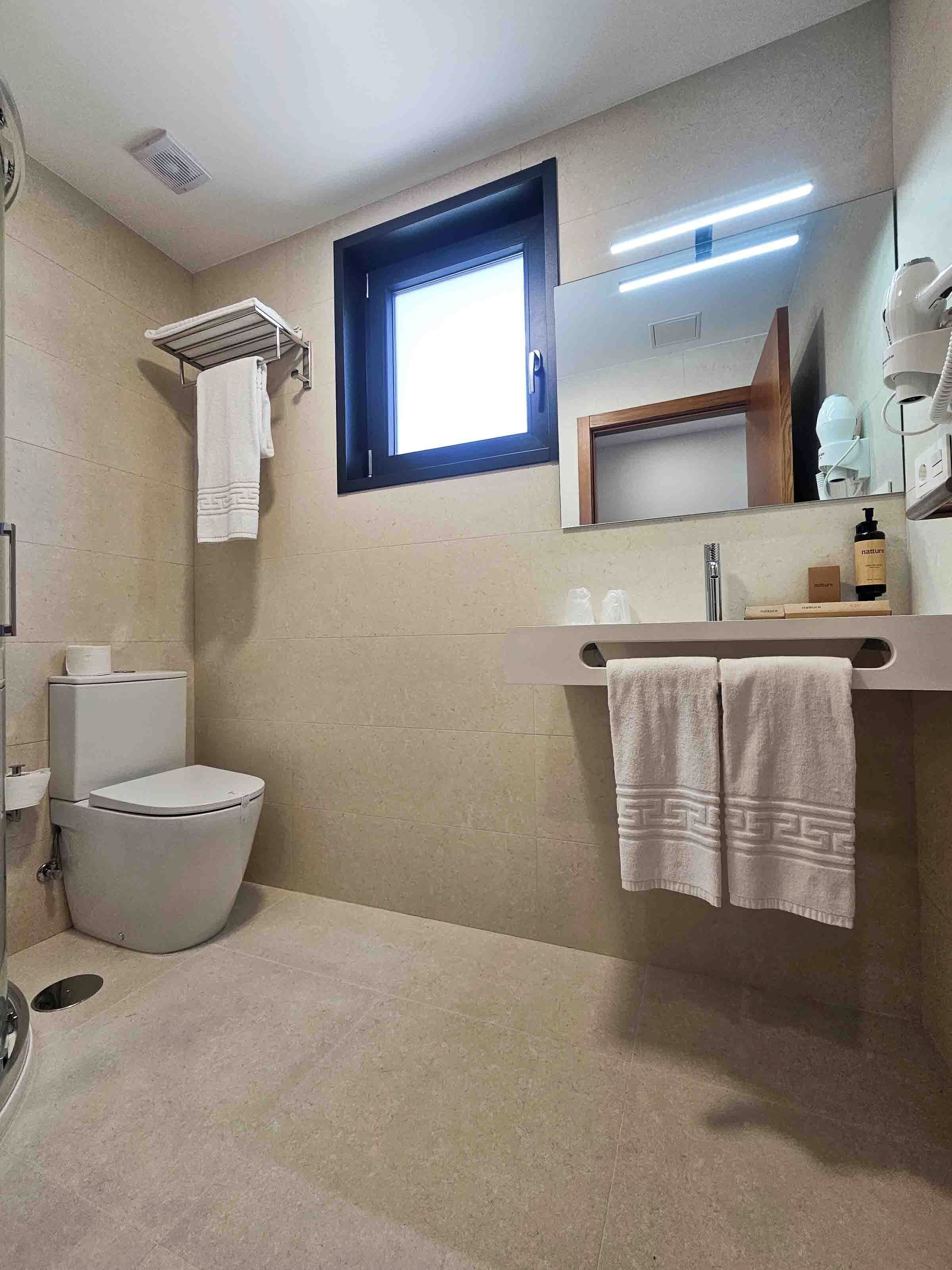 Uma casa de banho moderna com sanita, lavatório e espelho. Há uma janela sobre o lavatório e um porta-toalhas com toalhas.