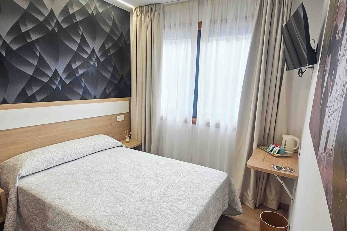 Quarto de hotel com cama, escrivaninha, televisão e janela com cortinas.