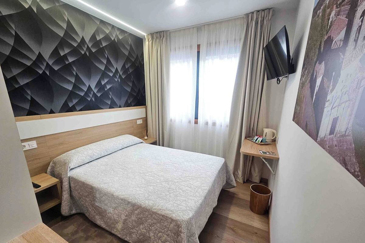 Quarto de hotel com cama, cabeceira de madeira, escrivaninha com cadeira, televisão na parede e janela com cortinas.