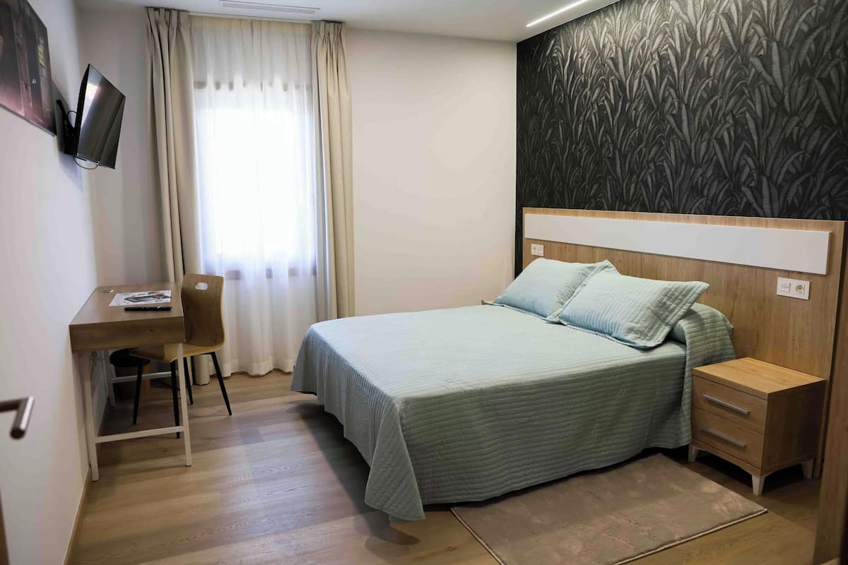 Quarto com cama, mesa de cabeceira, cadeira, escrivaninha e televisão fixada na parede.