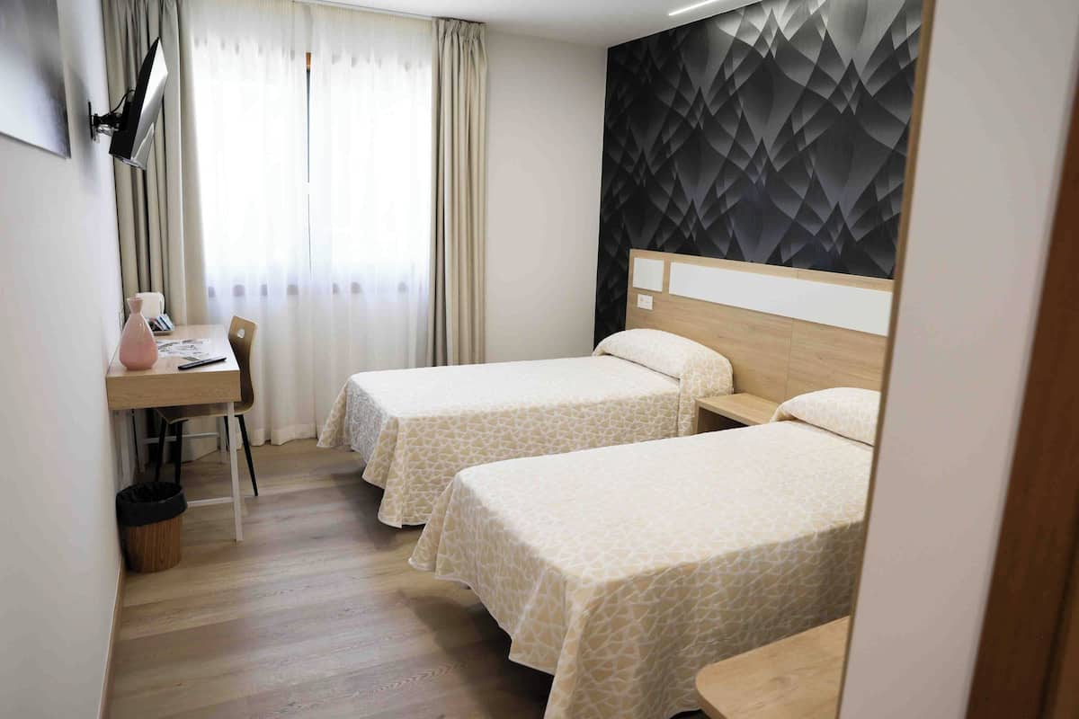 Quarto de hotel com duas camas, uma mesa com cadeira, um cesto de lixo e uma janela com cortinas.