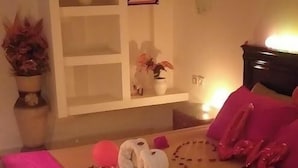 Quarto com cama, almofadas rosa, mesa de cabeceira, prateleira com objetos decorativos e criado-mudo com abajur.