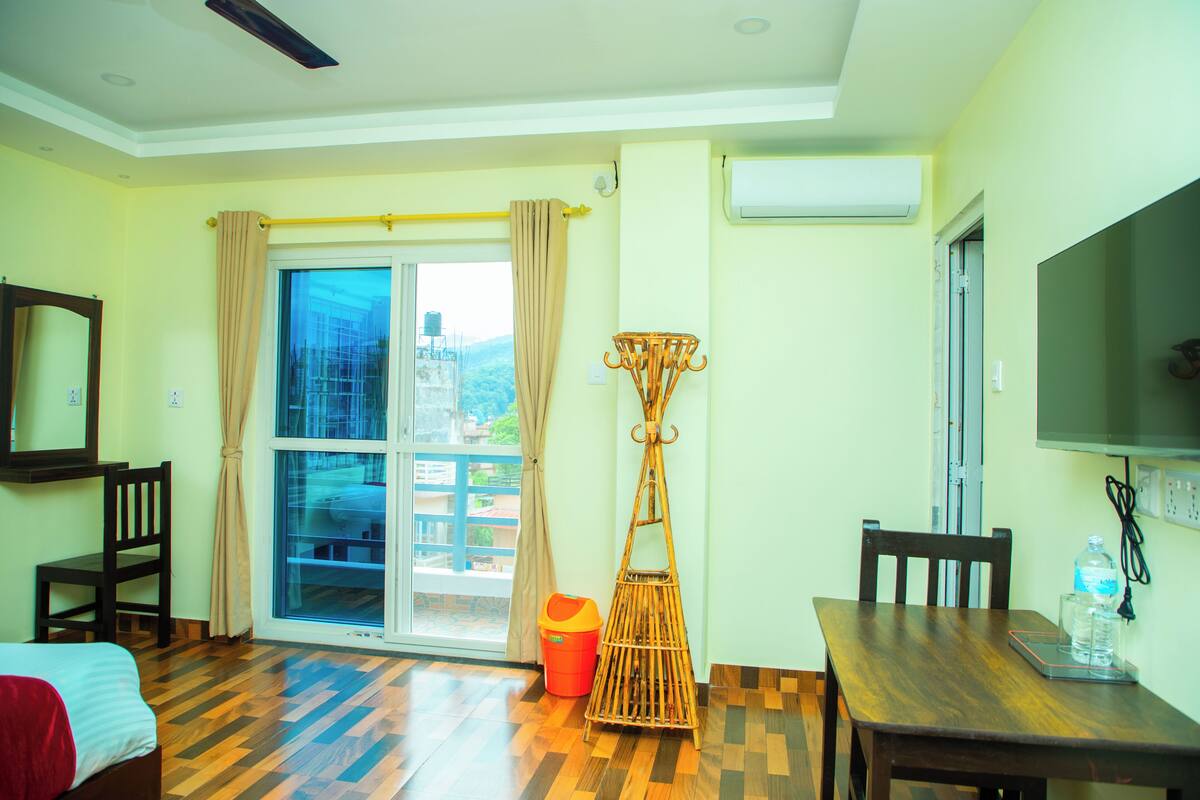Quarto de hotel com cama, mesa de jantar, televisão, janela com cortinas e vista para o exterior.