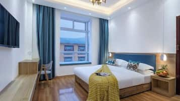 Quarto de hotel com cama, escrivaninha, cadeira, janela com cortinas, mesa de cabeceira com abajur e vista para um edifício.