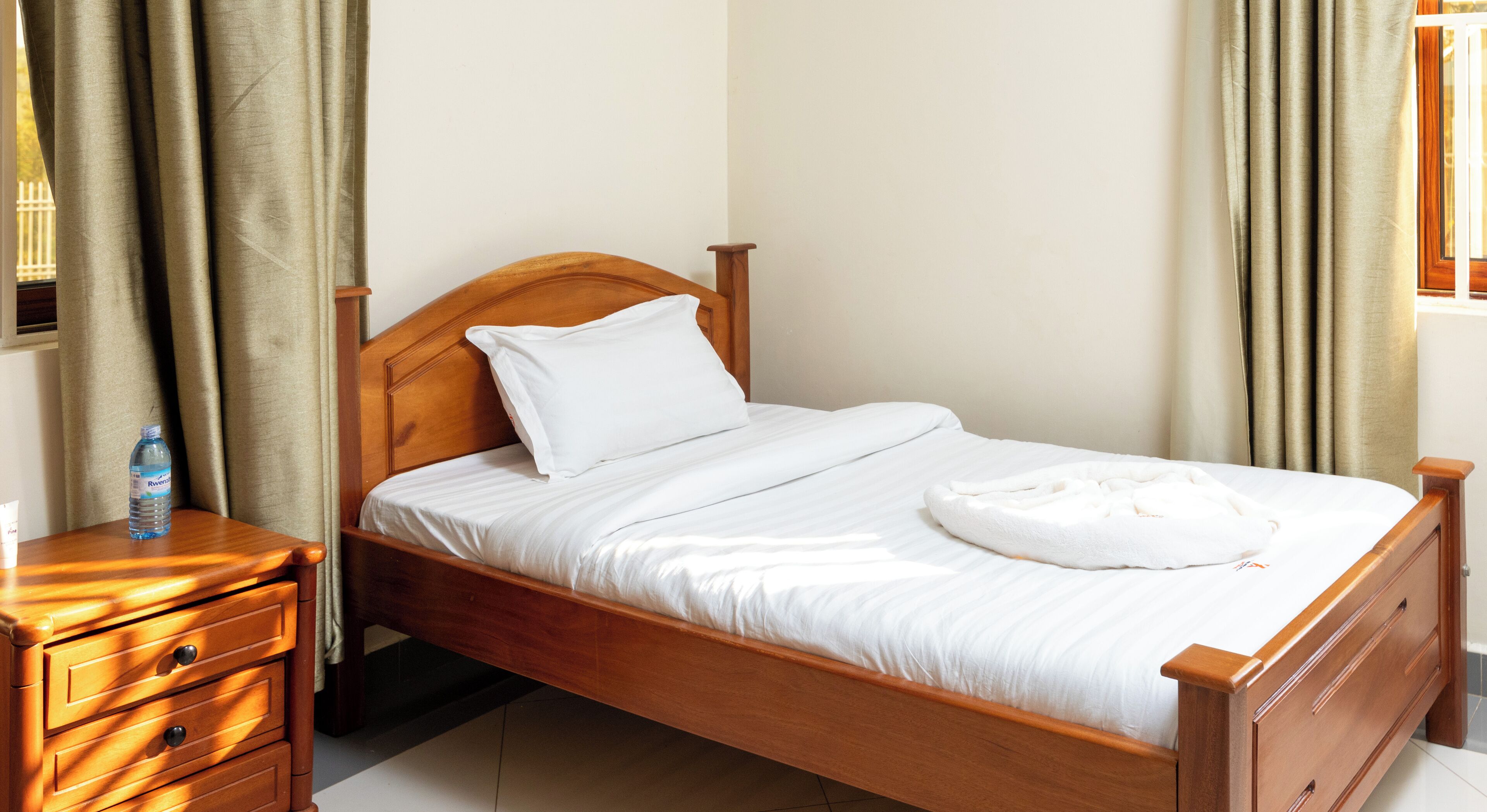 Uma cama de madeira com roupa de cama branca, um criado-mudo de madeira com gavetas e uma garrafa de água.